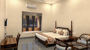 Quarto de hotel com cama, escrivaninha, cadeira, janela com cortinas, ar condicionado e ventoinha no teto.