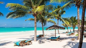 Uma praia com palmeiras, espreguiçadeiras e um guarda-sol de palha.