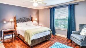 Quarto com cama, duas mesas de cabeceira com luminárias, uma cadeira, janela com cortinas e ventilador de teto.