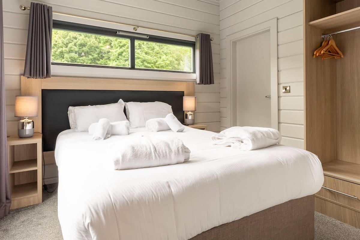 Cama bem arrumada com lençóis e toalhas brancas, um abajur e uma janela com cortinas.