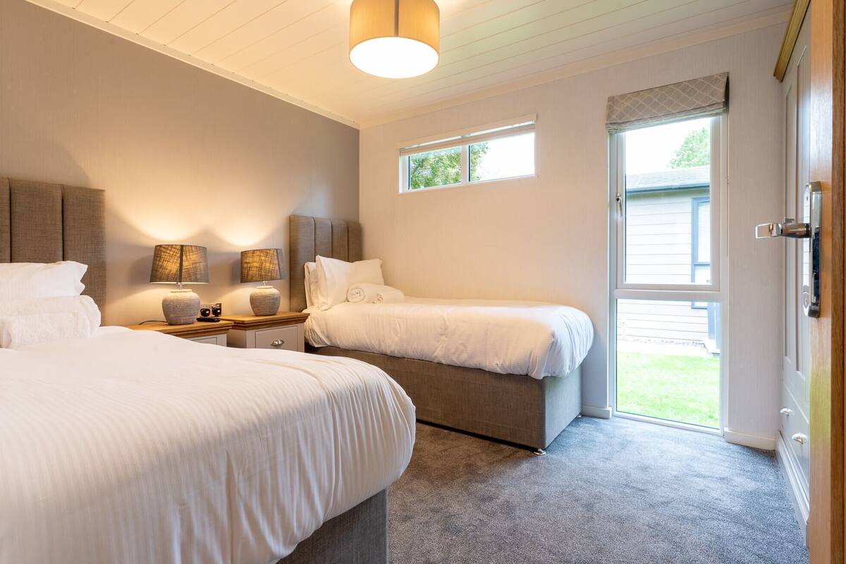 Quarto moderno com duas camas, uma mesa de cabeceira, um abajur e uma janela com vista para o exterior.