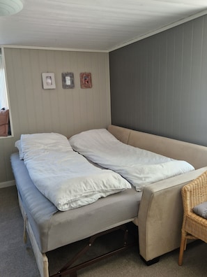 Uma cama de solteiro com colchão e almofadas, uma mesa pequena e uma cadeira num quarto com painéis de madeira e parede cinzenta.