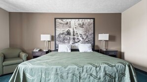Quarto de hotel com uma cama grande, duas mesas de cabeceira com abajures, uma cadeira e um quadro pendurado na parede com uma cascata.