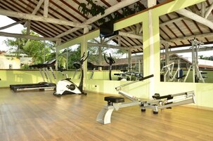 Academia com máquinas de cardio e equipamentos de peso, piso de madeira e área coberta com vista para o exterior.