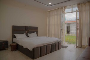 Quarto com uma cama grande, uma mesinha de cabeceira, um cesto de lixo e vista para um jardim através de uma porta aberta.