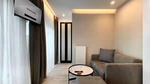 Uma sala de estar moderna com um sofá, um banquinho e uma mesa pequena com um controle remoto. Uma televisão está montada na parede, e há um radiador no canto.
