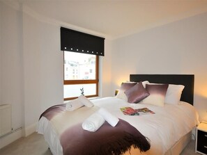 Cama bem arrumada com cabeceira escura, roupa de cama branca e janela com vista para edifícios.