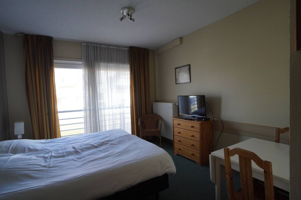 Quarto de hotel com cama, escrivaninha, cadeira, televisão e janela com cortinas.