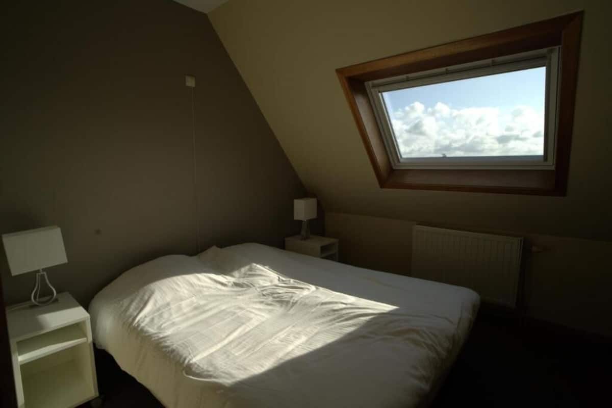 Quarto com uma cama de solteiro, mesa de cabeceira, abajur, janela com vista para o céu e radiador.