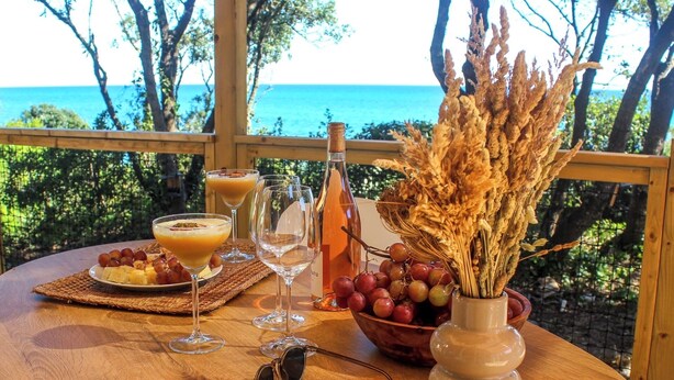 Mesa posta com garrafa, copos, taça de uvas e prato de frutas, com vista para a costa.