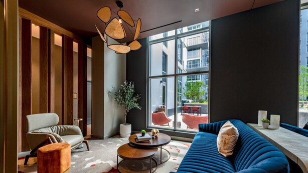 Uma área de estar moderna com sofá, poltrona e mesa de centro redonda. Grandes janelas oferecem vista para o exterior.