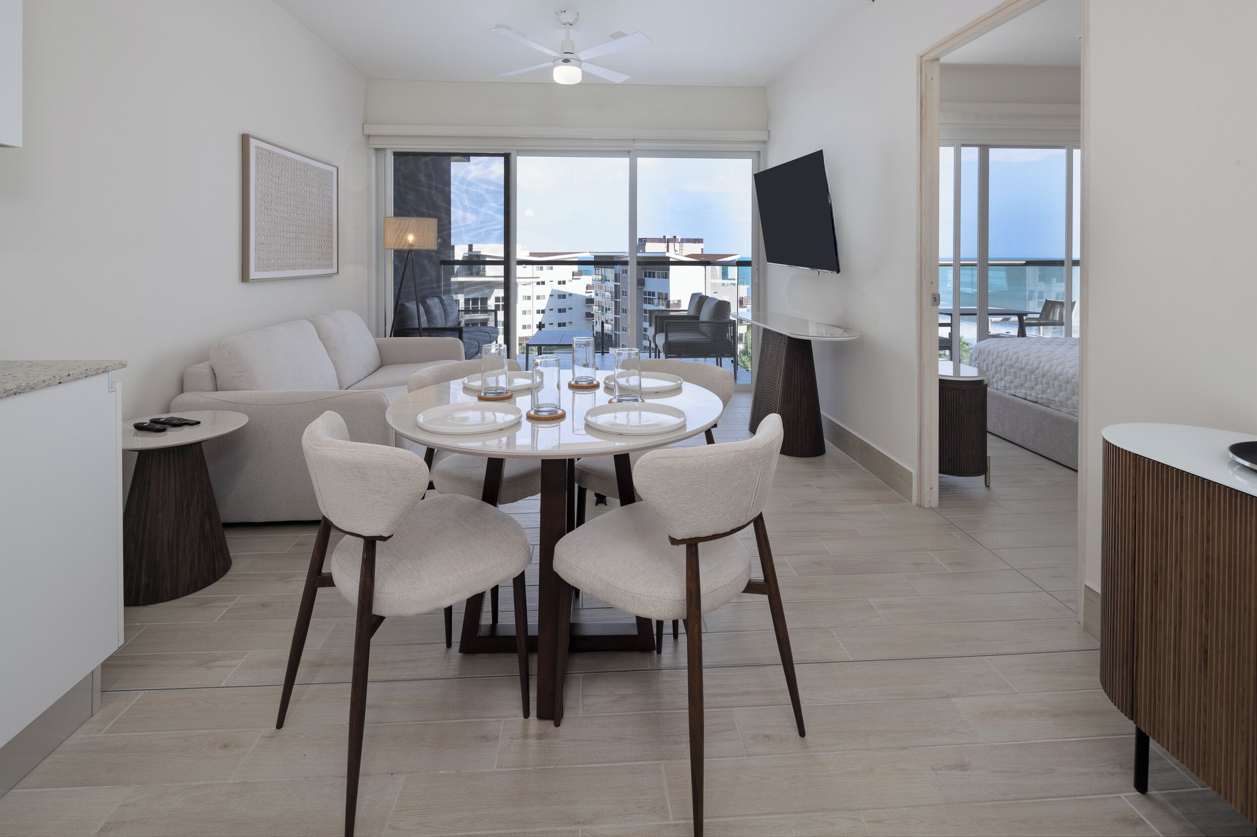 Uma sala de estar moderna com uma mesa de jantar redonda, quatro cadeiras, um sofá, uma TV e uma varanda com vista para a cidade.