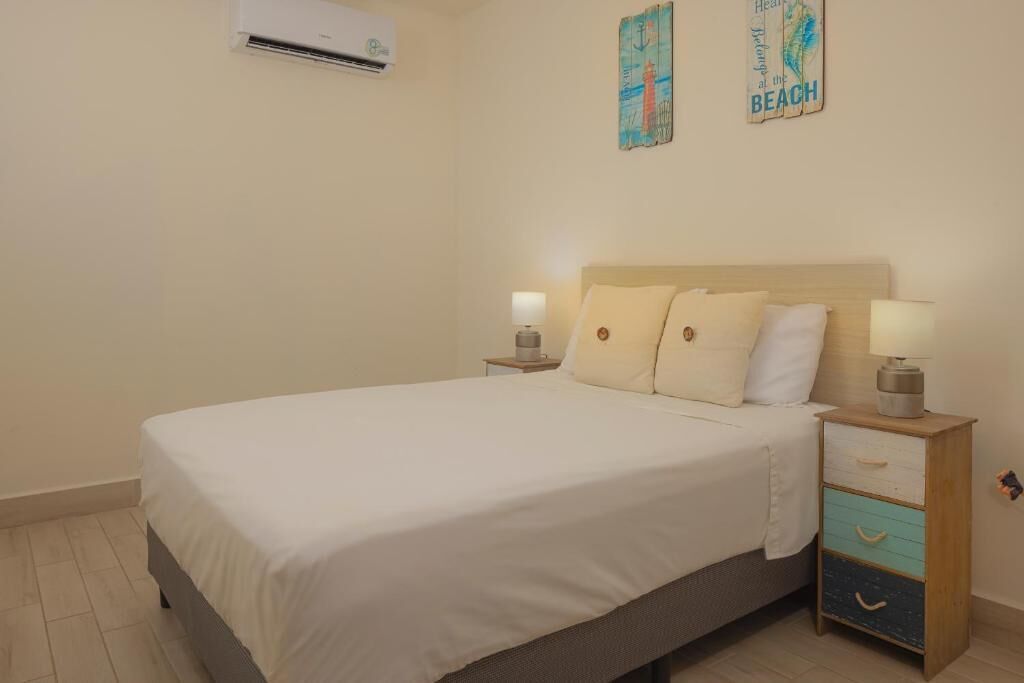 Quarto com cama, duas mesas de cabeceira com luminárias, ar condicionado fixado na parede e dois objetos decorativos na parede.