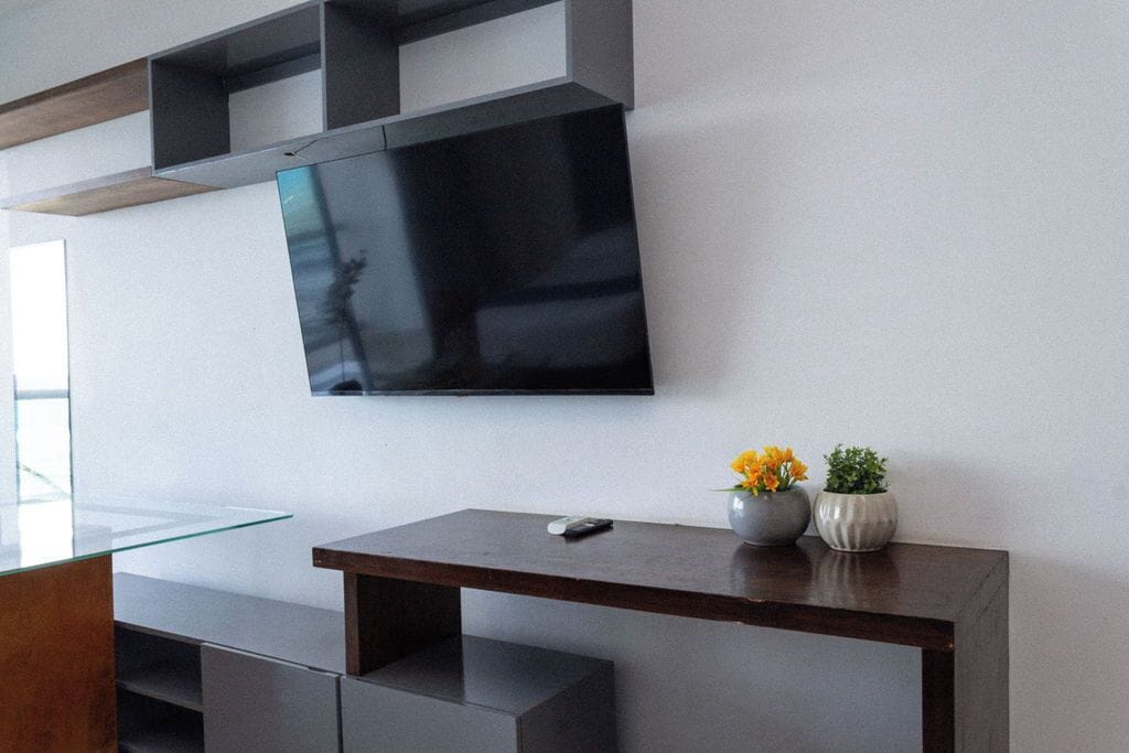 Uma sala de estar moderna com uma TV de tela plana na parede, uma mesa de centro de vidro e um aparador de madeira escura com um vaso de flores.