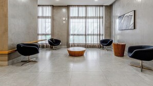 Uma área de espera moderna com uma mesa de centro redonda de madeira, quatro cadeiras pretas e uma janela grande com cortinas translúcidas.
