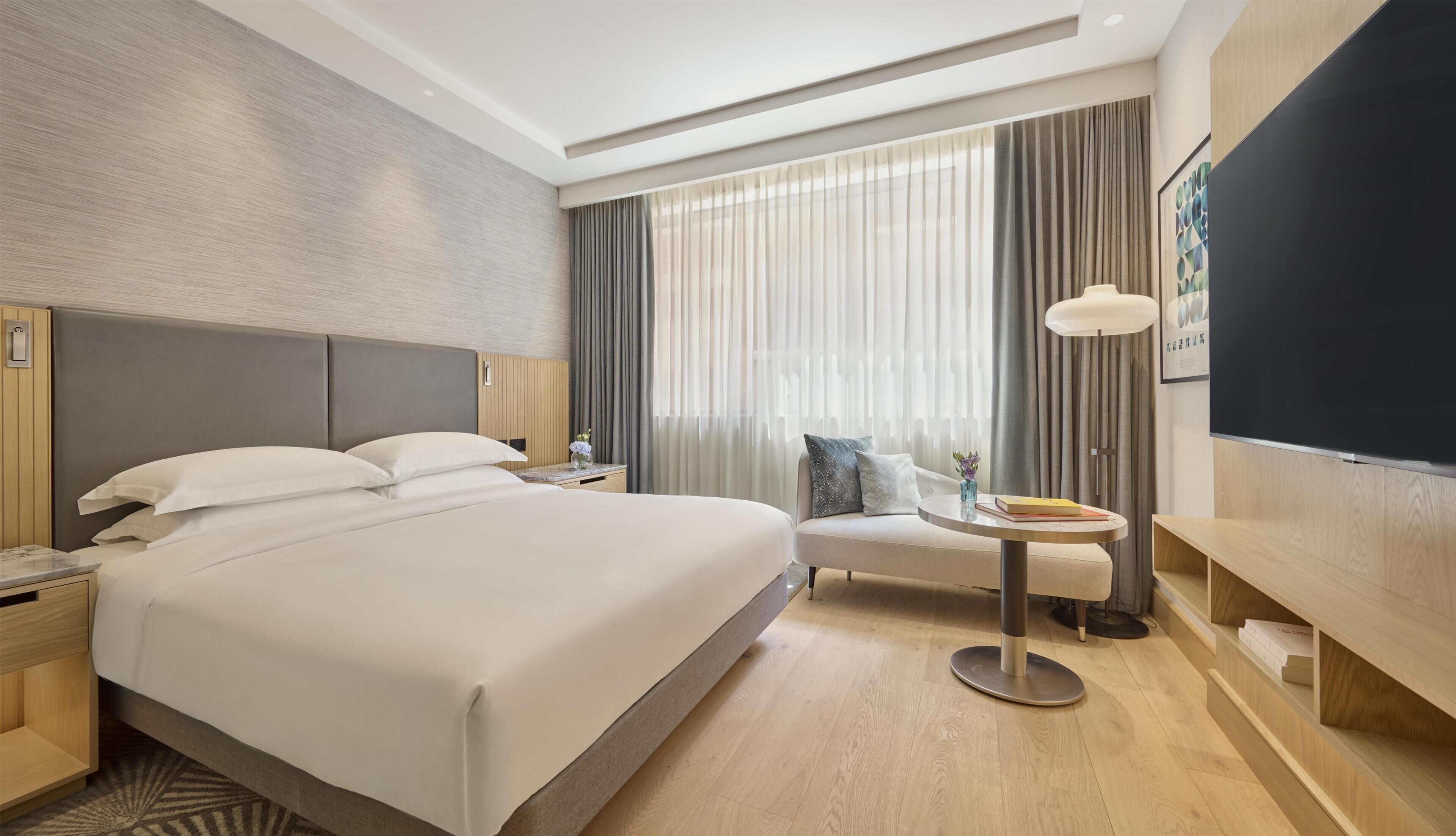 Quarto de hotel com uma cama grande, mesa de cabeceira, mesa redonda pequena, cadeira, abajur, televisão e janela com cortinas.