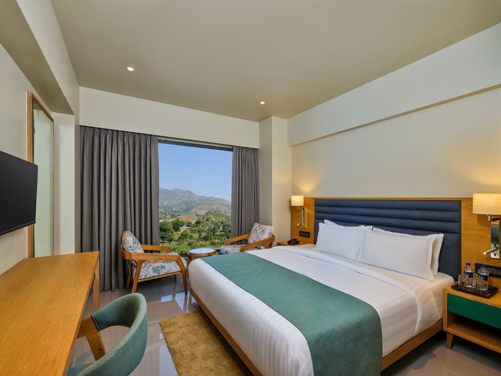 Quarto de hotel com uma cama grande, escrivaninha, cadeira, televisão e vista para as montanhas.