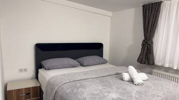 Uma cama bem arrumada com cabeceira, uma mesa de cabeceira com gavetas e uma janela com cortinas.