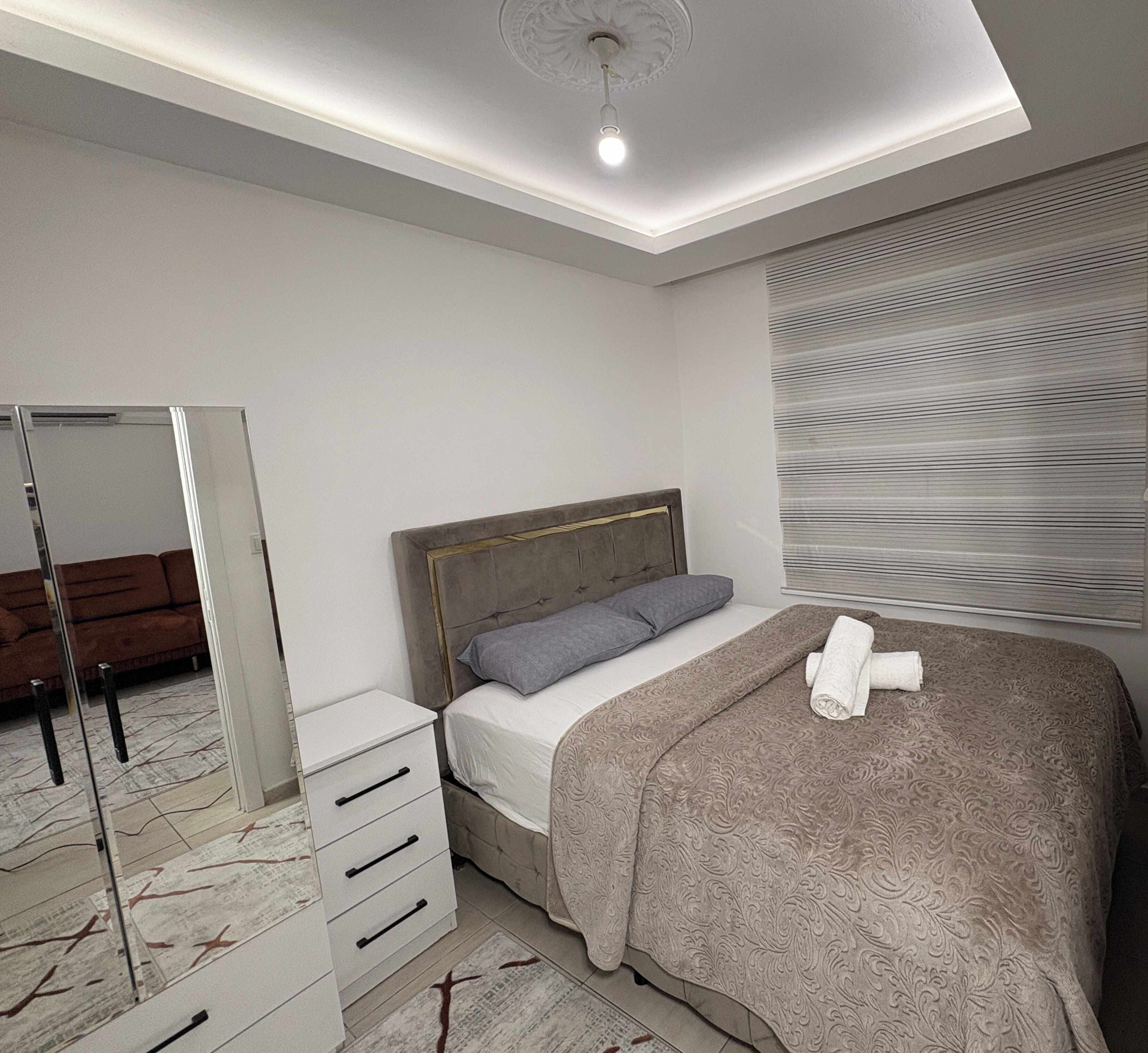 Quarto com cama, mesa de cabeceira, penteadeira, espelho e luminária no teto.