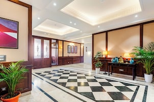 Um saguão de hotel com piso xadrez, painéis de madeira e uma recepção com plantas em vasos.