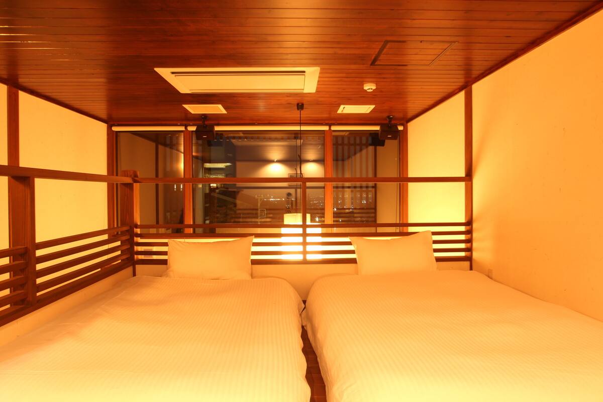Quarto com duas camas, teto de madeira e janelas amplas.