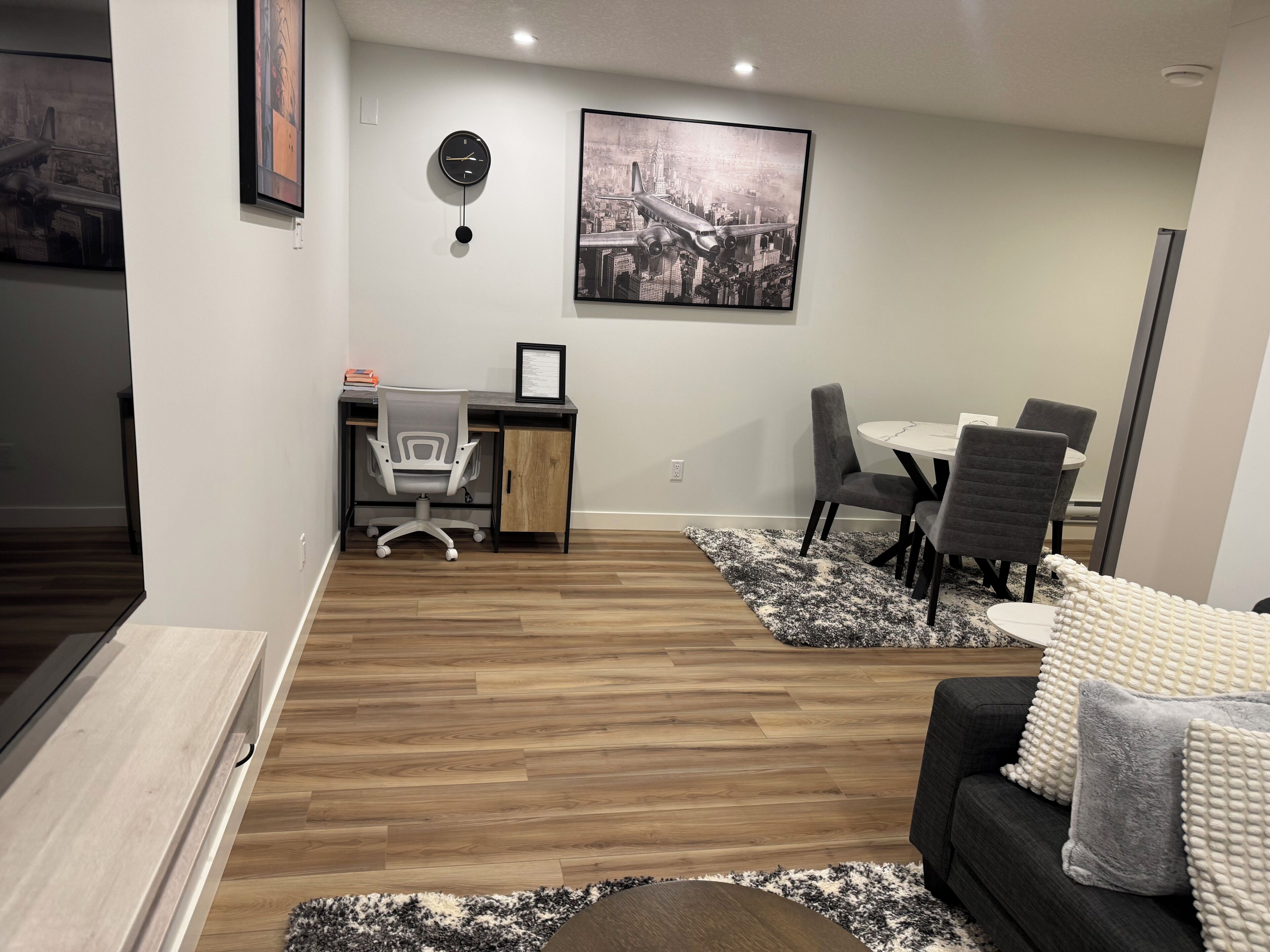 Uma sala de estar moderna com assoalho de madeira escura, um sofá cinza, uma área de jantar com mesa branca e cadeiras cinzas, uma cadeira de escritório cinza e um relógio preto na parede.