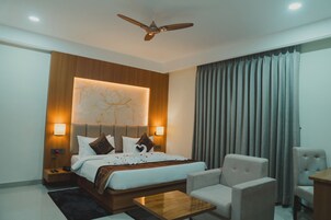 Quarto de hotel moderno com uma cama grande, um ventilador de teto, uma luminária na parede, uma mesa de apoio e um espaço de estar com uma cadeira e uma mesinha.