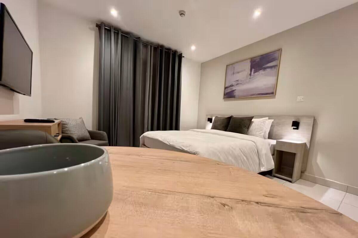 Quarto de hotel moderno com uma grande mesa de madeira, uma cama com roupa de cama branca, um sofá cinzento e um quadro na parede.