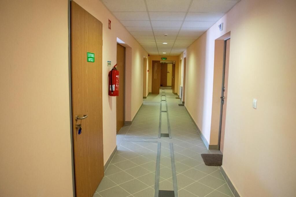 Um corredor comprido e estreito com portas em ambos os lados, um extintor fixado na parede e uma placa verde indicando um elevador.