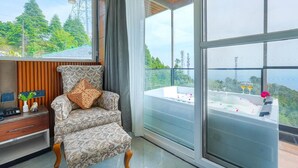 Quarto com poltrona estampada, mesa lateral de madeira e grande janela com vista para uma varanda com hidromassagem e vegetação.
