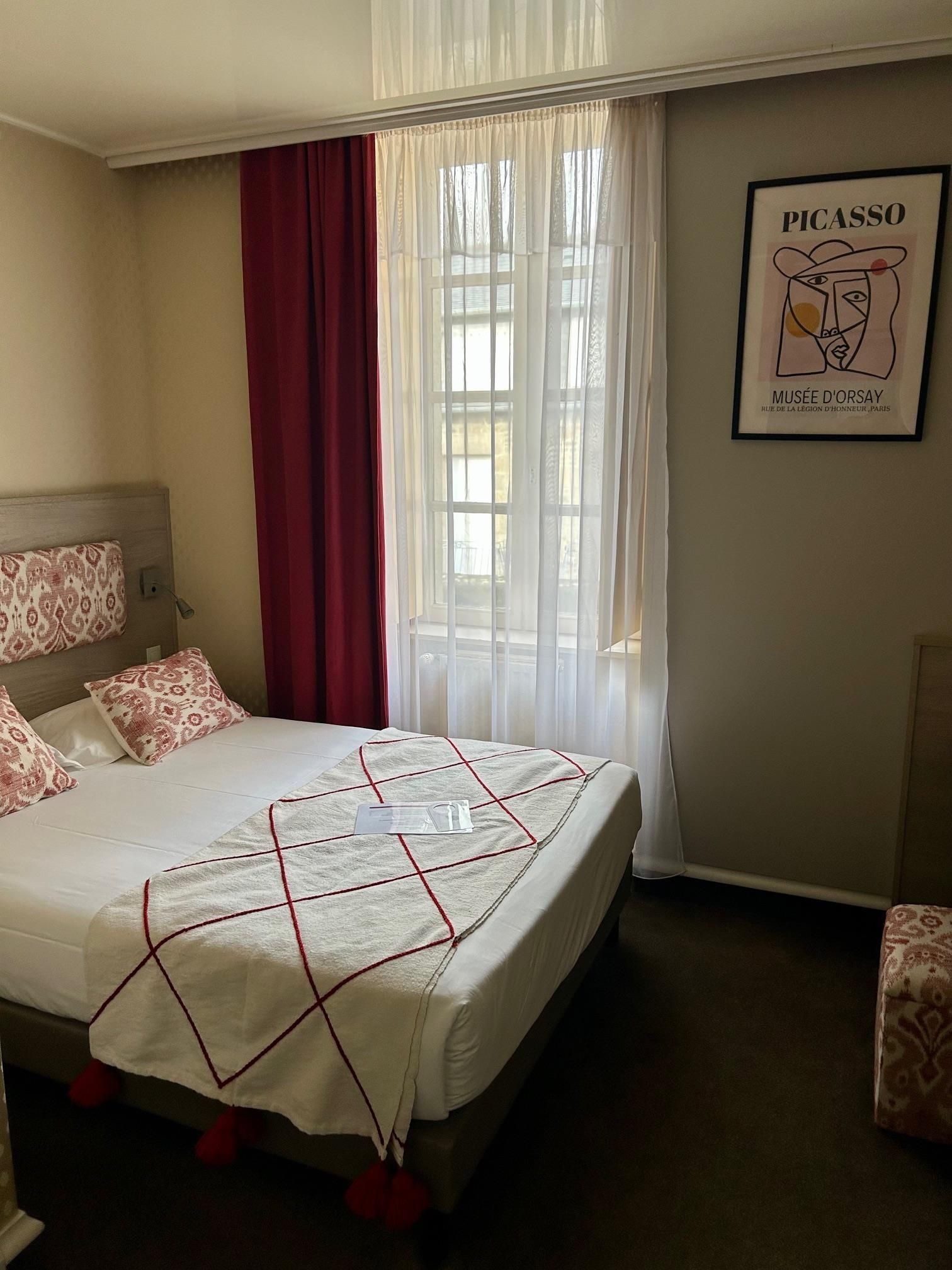 Quarto de hotel com cama, janela com cortinas finas, cortina vermelha, obra de arte de Picasso emoldurada e almofadas estampadas.