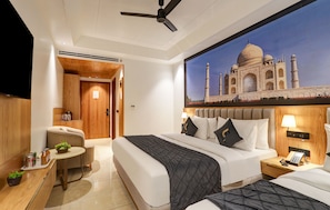 Quarto de hotel com uma cama, uma escrivaninha, uma cadeira, uma televisão e um mural na parede com vista para o Taj Mahal.