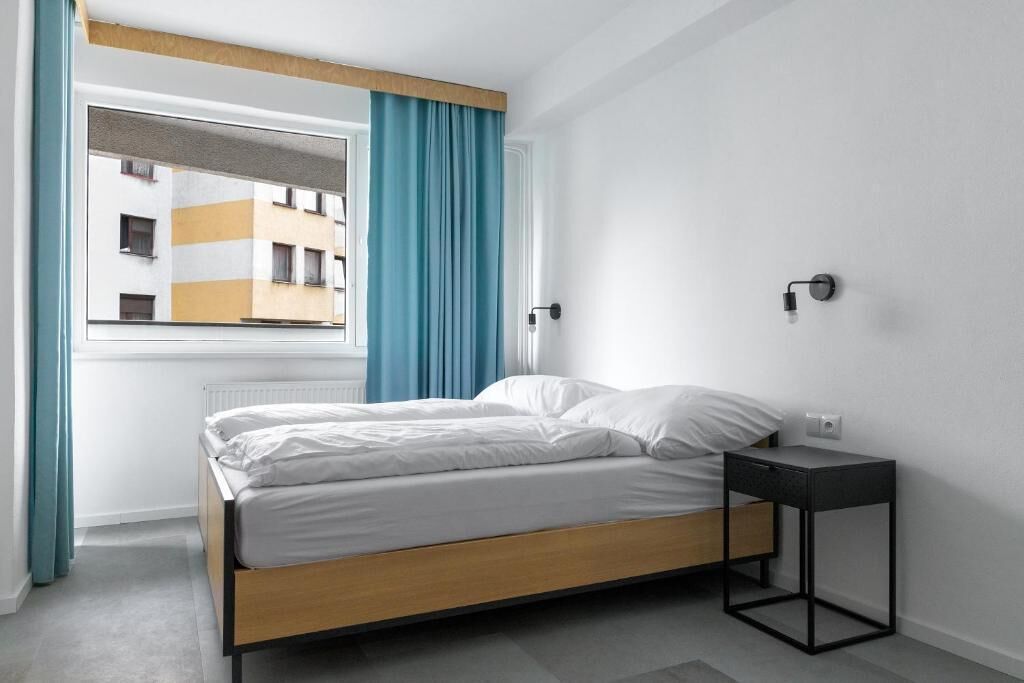 Quarto moderno com cama, mesa de cabeceira, janela com cortinas e vista para edifícios.