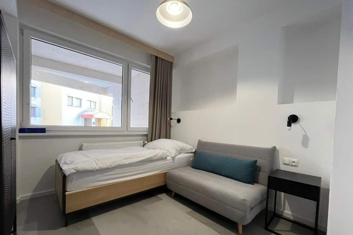 Quarto pequeno e moderno com cama, sofá, mesa de cabeceira e janela.