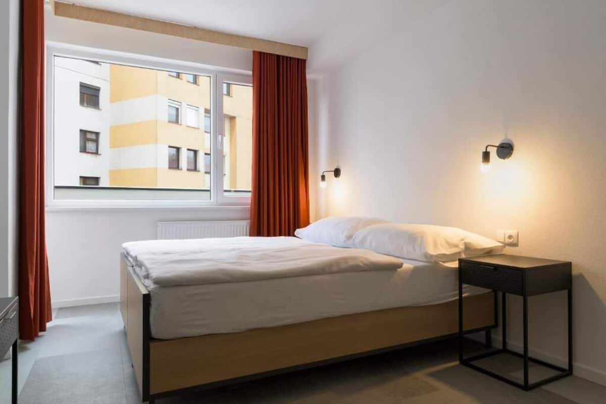 Quarto com cama, mesa de cabeceira, janela com cortinas e luminária na parede.