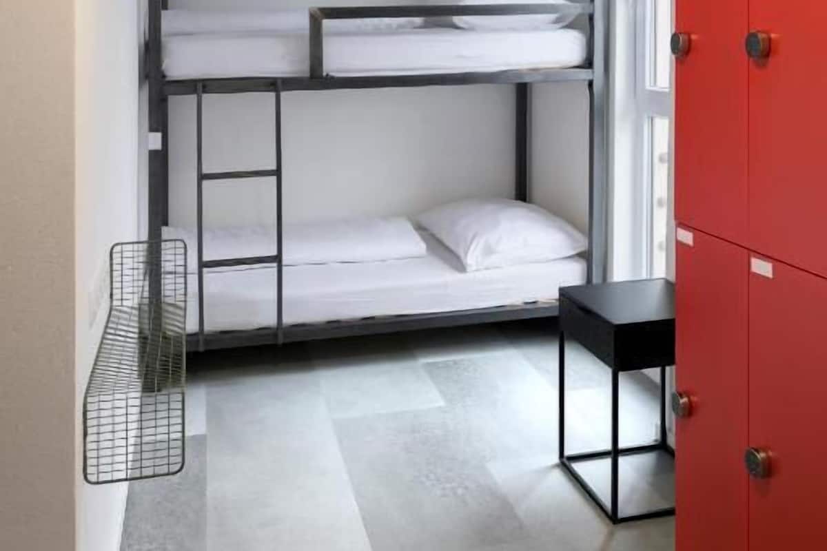 Uma cama de solteiro com roupa de cama branca, um guarda-roupa vermelho e uma pequena mesa preta.