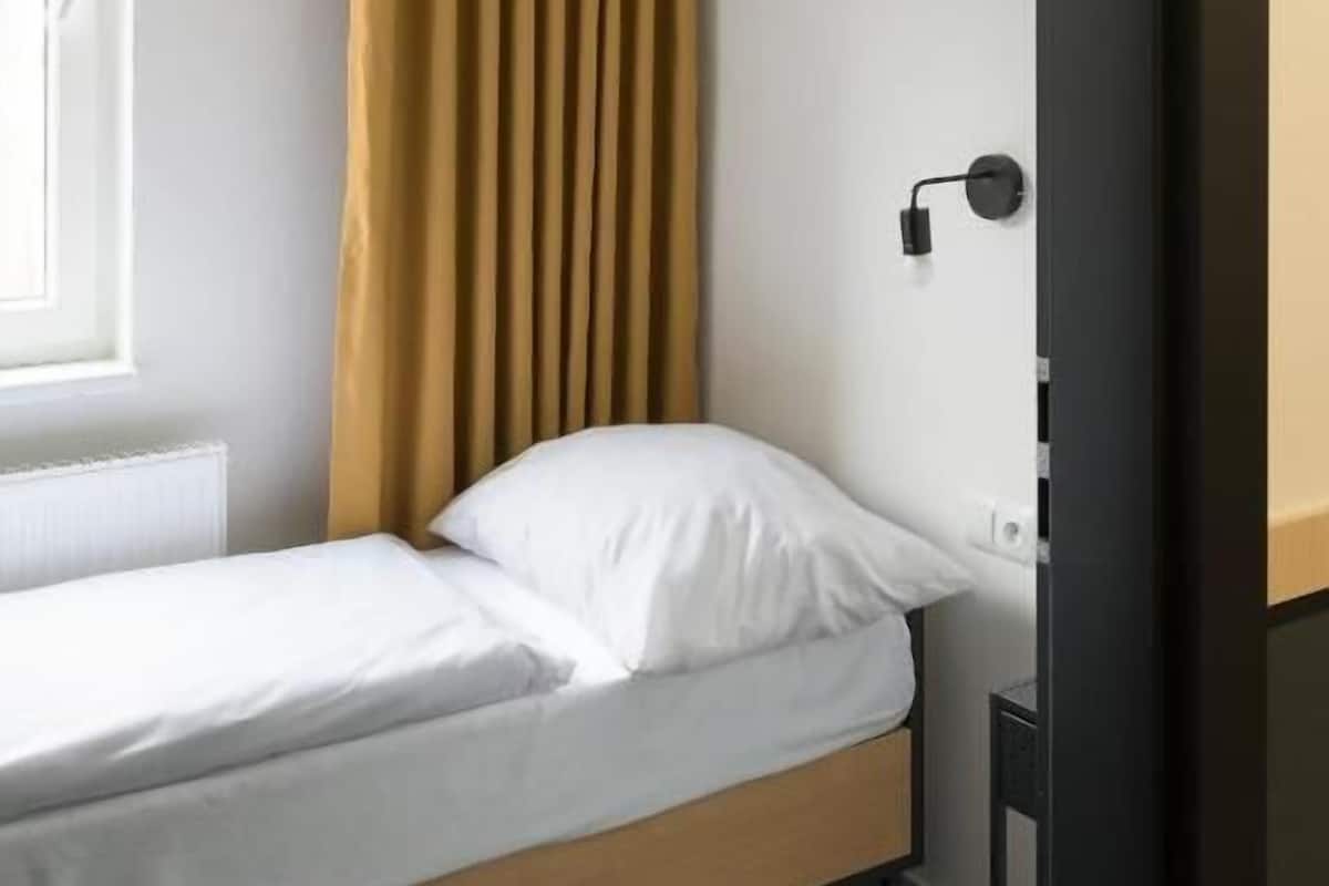 Uma cama de solteiro com estrutura de madeira, colchão branco e luminária pendente amarela.