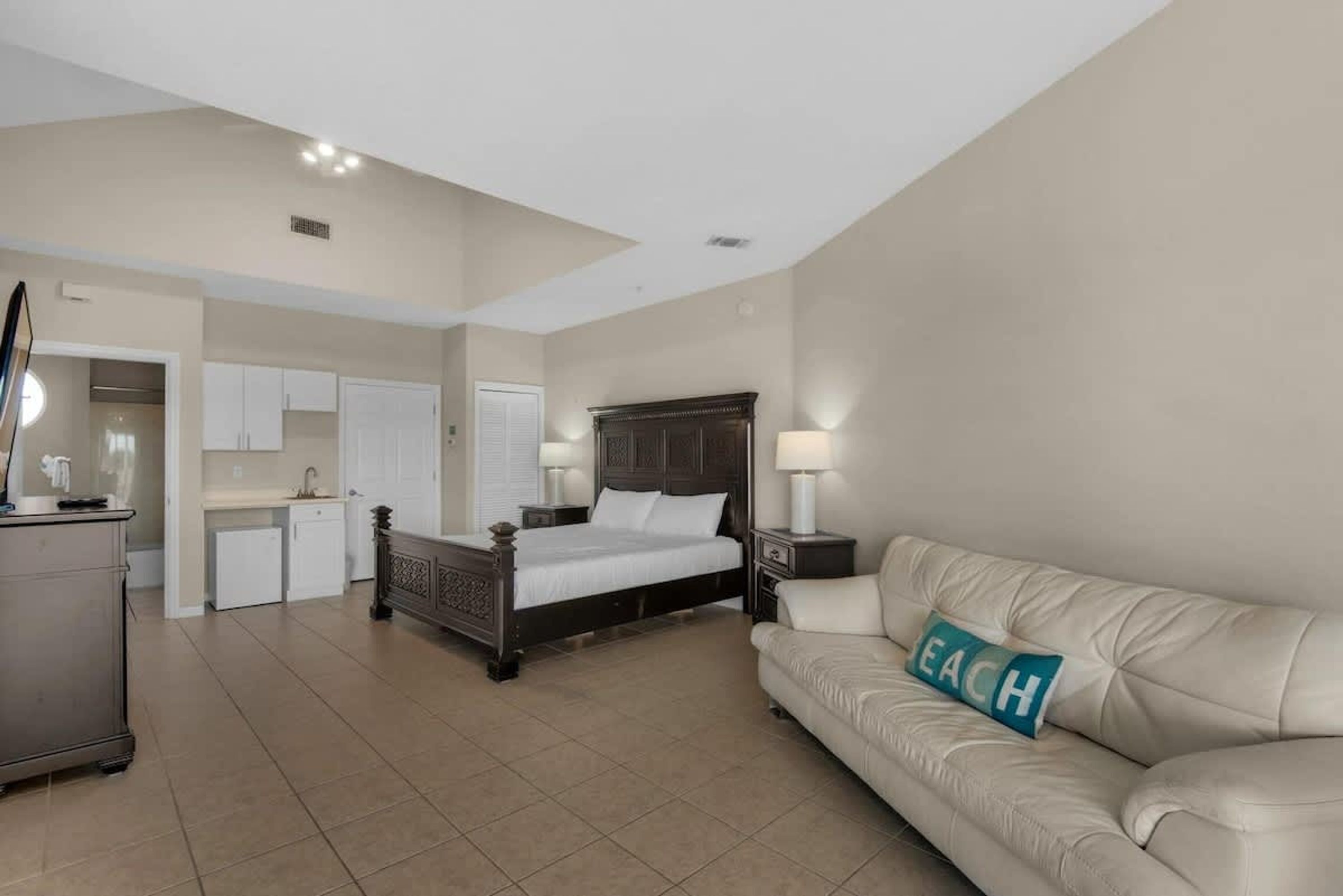 Quarto de hotel com cama, sofá, escrivaninha e cozinha compacta.