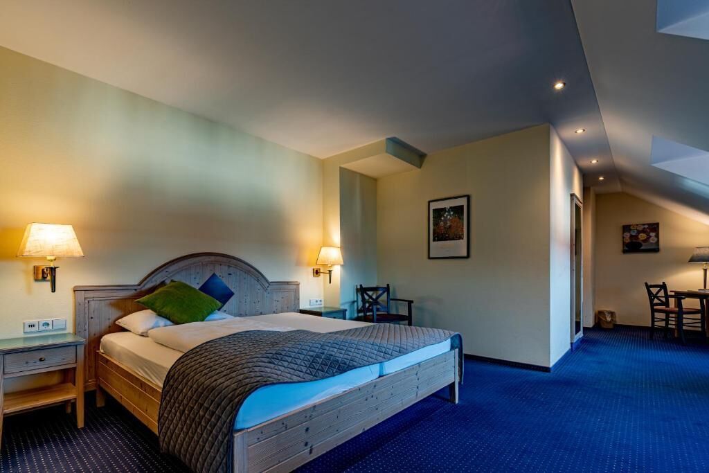 Quarto de hotel com uma cama grande, duas mesas de cabeceira com abajures, uma cadeira e uma mesinha.