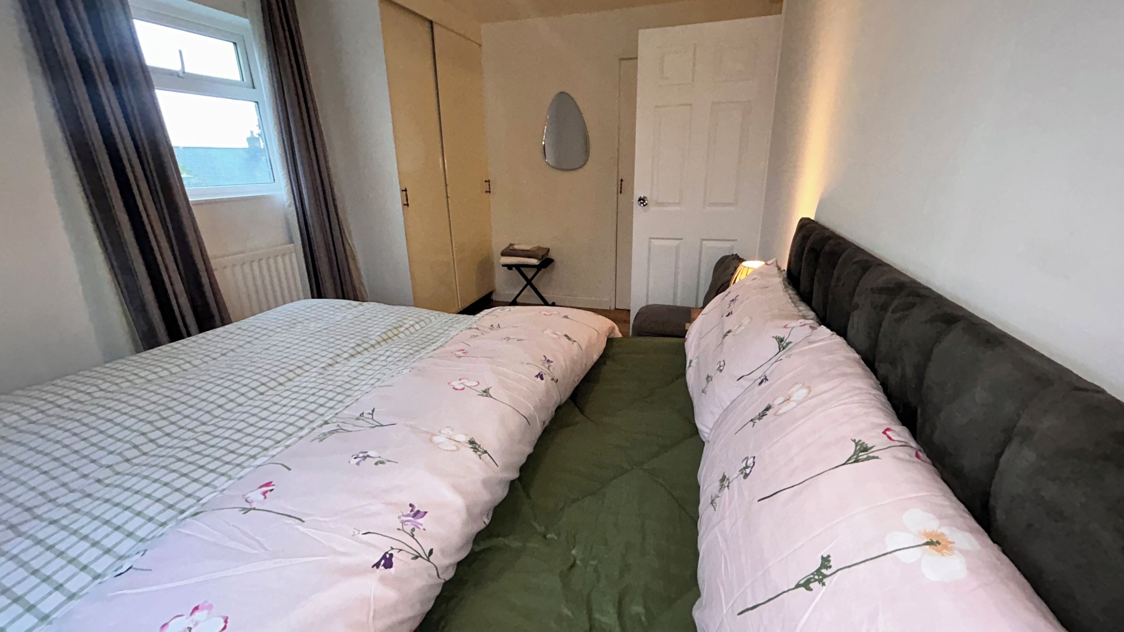 Quarto com duas camas, janela com cortinas, porta e pequena mesa com abajur.