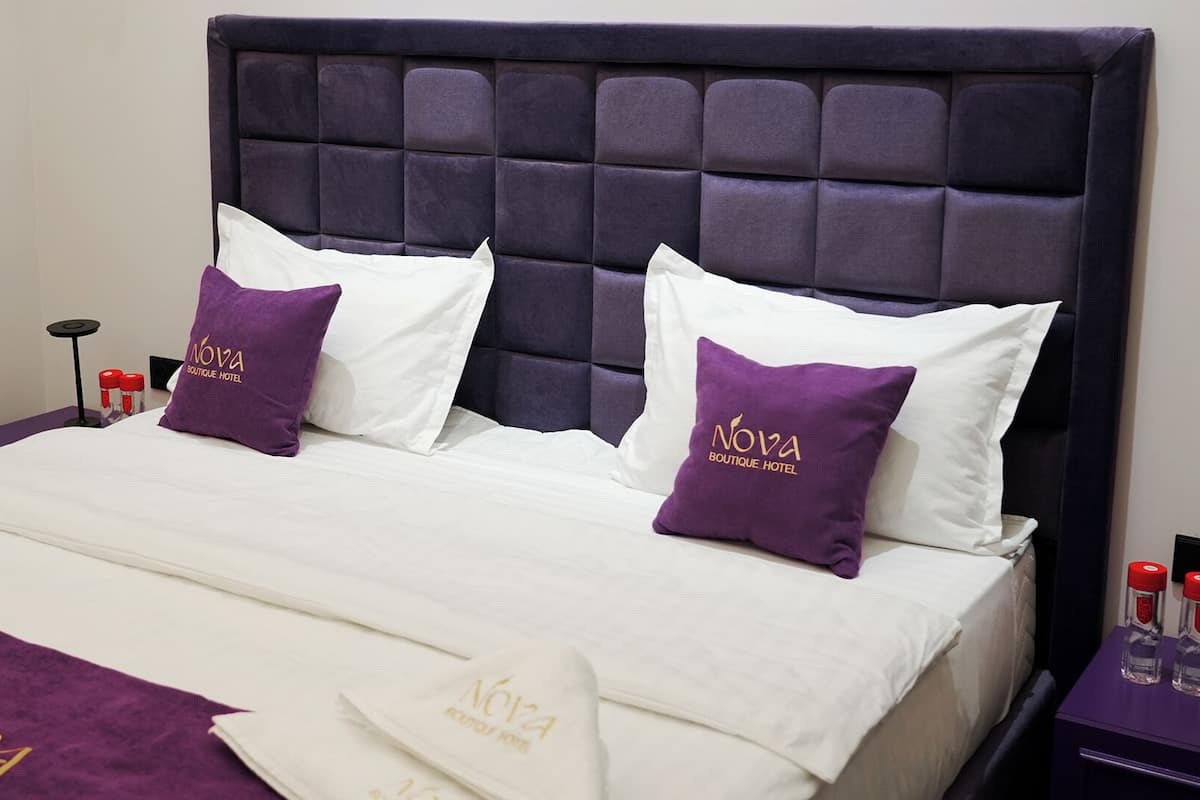 Quarto de hotel com uma cama grande, duas almofadas com o logotipo do hotel, um cobertor roxo e uma mesa de cabeceira com uma luminária e garrafas.