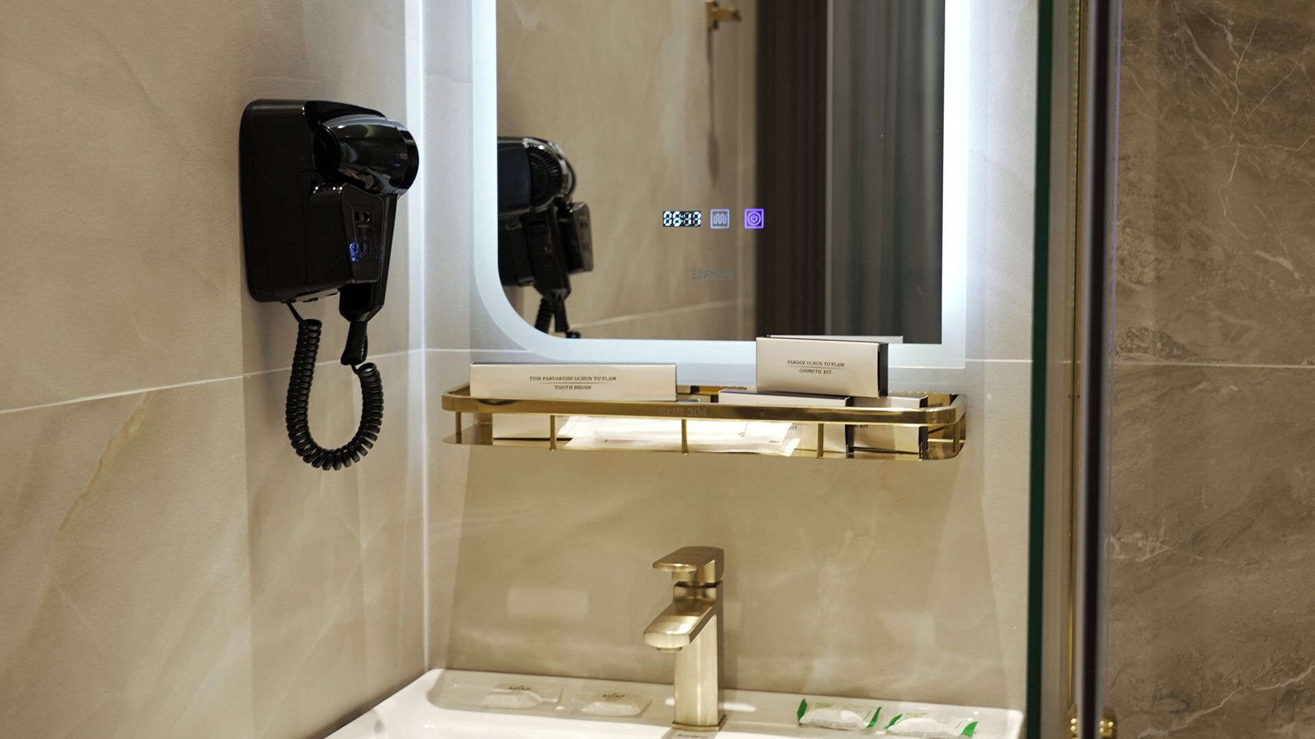 Um banheiro com telefone fixado na parede, espelho, pia com torneira dourada e prateleira com necessaire.