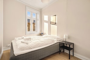 Uma cama bem arrumada com lençóis brancos, uma mesa de cabeceira com abajur e uma janela com persianas.