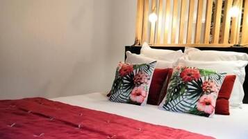 Cama bem arrumada com manta vermelha e almofadas florais, cabeceira de madeira e luminária fixada na parede.