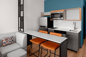 Cozinha compacta com micro-ondas, frigorífico e máquina de lavar louça. Dispõe de um espaço de refeições com uma mesa e duas cadeiras.