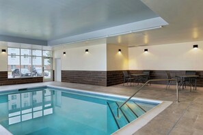 Uma piscina com um deck revestido, área de estar com mesas e cadeiras, e vista para o exterior através de grandes janelas.