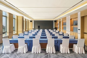 Uma sala de conferências com mesas e cadeiras dispostas em fileiras, janelas amplas e decoração moderna.