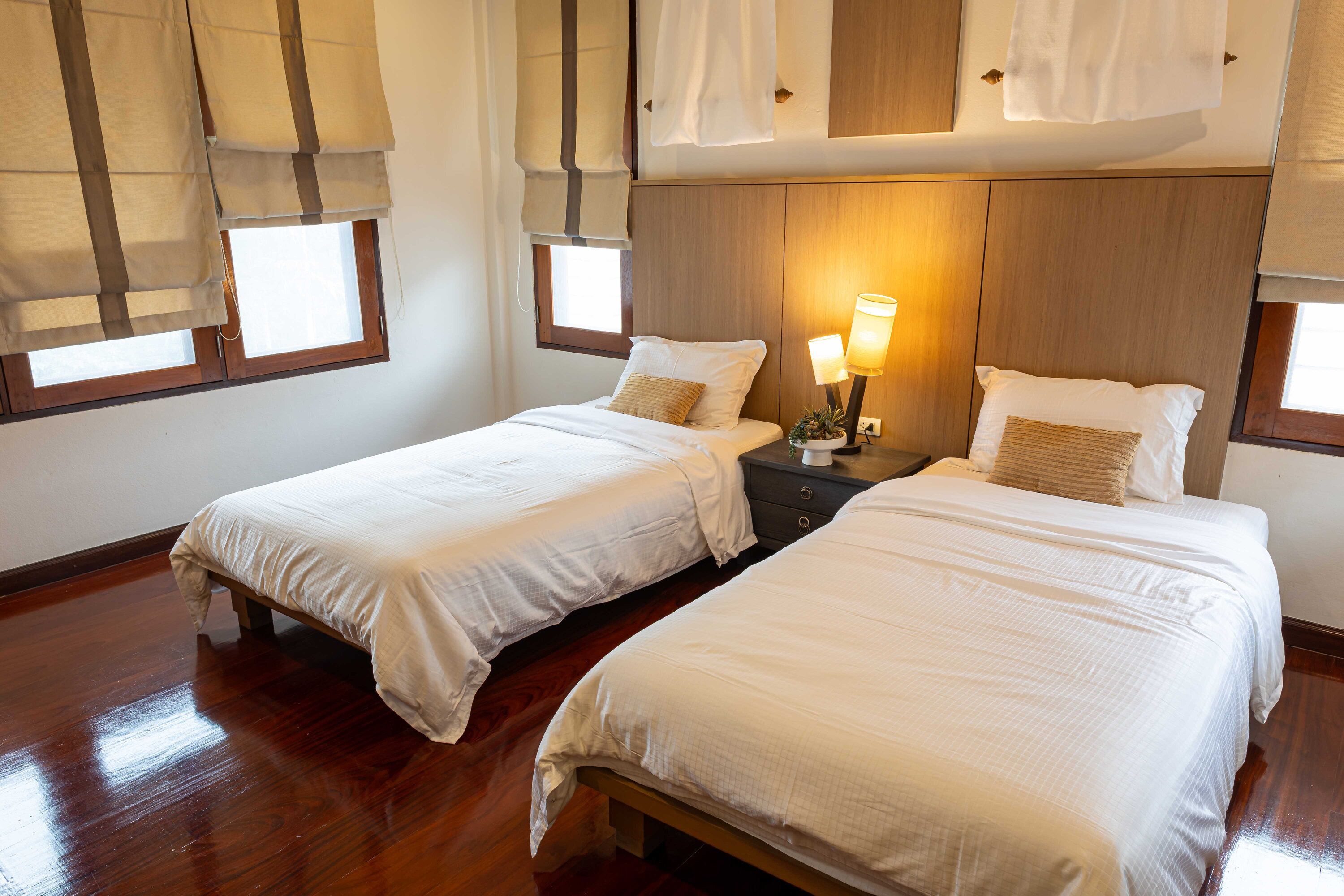 Quarto de hotel com duas camas de solteiro, piso de madeira, mesa de cabeceira com abajur e janela com cortinas.
