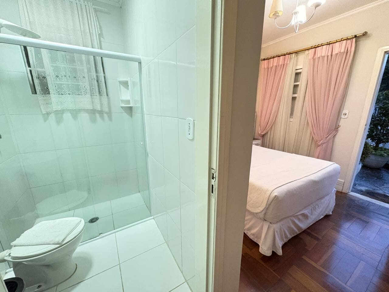 Um banheiro com box de vidro, vaso sanitário e cama com cortinas cor-de-rosa.