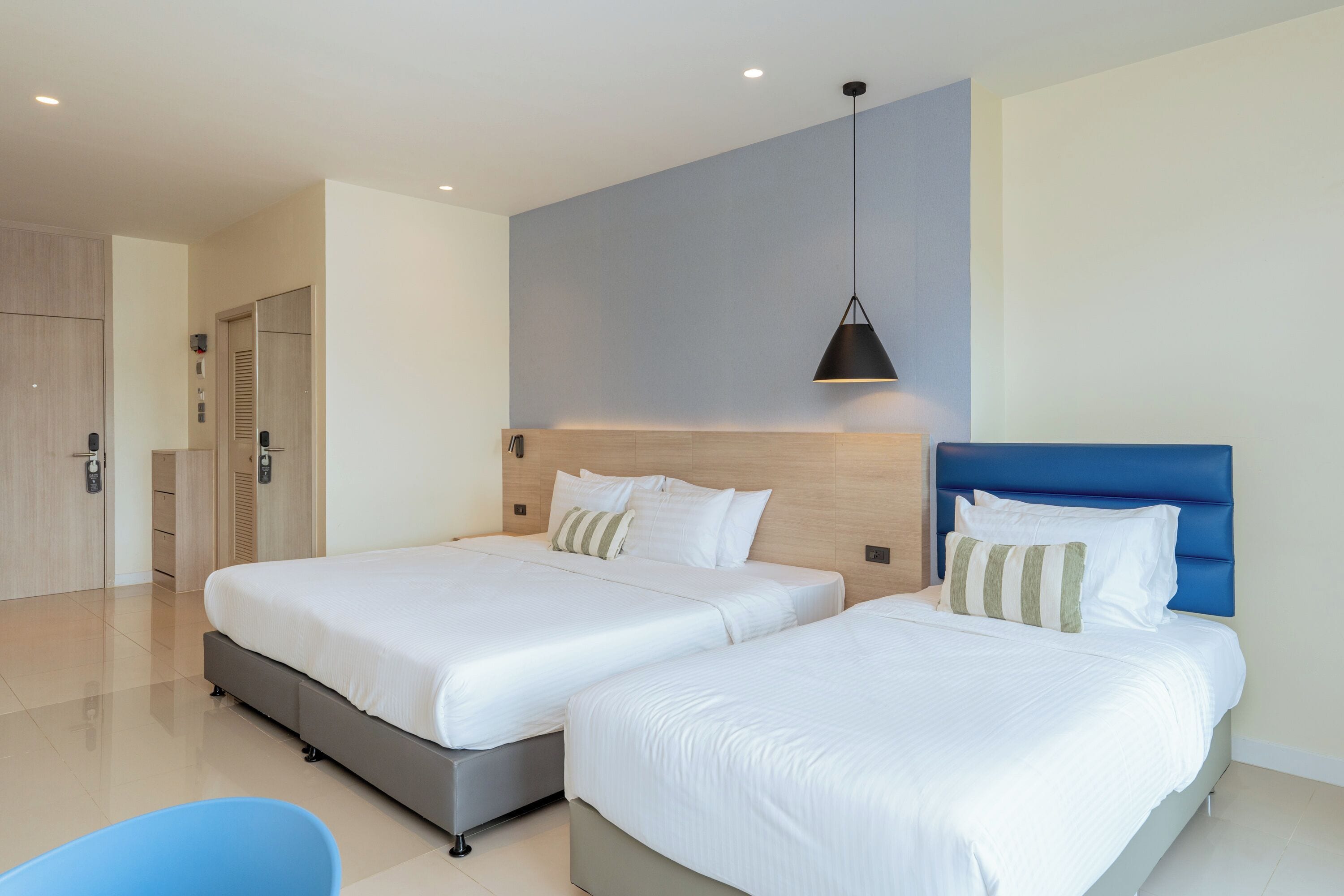 Quarto de hotel com duas camas, uma cadeira azul e uma luminária pendente moderna.